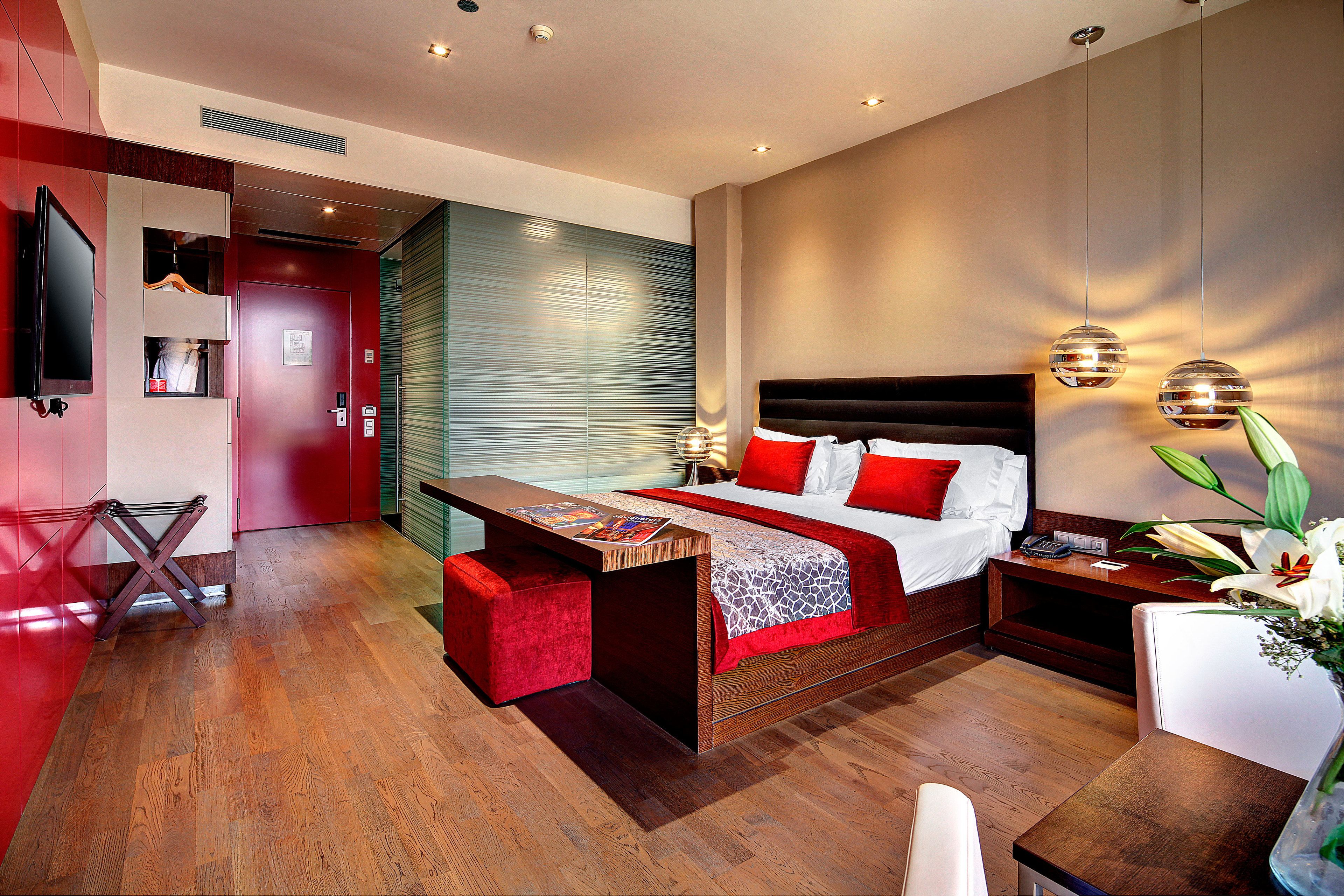 Quarto de hotel moderno com chão de madeira, cama com roupa de cama vermelha, escrivaninha e TV fixada na parede.