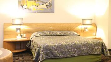 Quarto de hotel com cabeceira de madeira, edredom com padrão verde e cinza, duas luminárias de apoio e um quadro acima da cama.