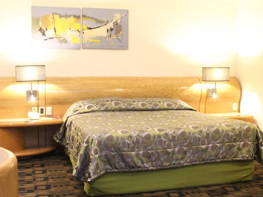 Quarto de hotel com cabeceira de madeira, edredom com padrão verde e cinza, duas luminárias de apoio e um quadro acima da cama.