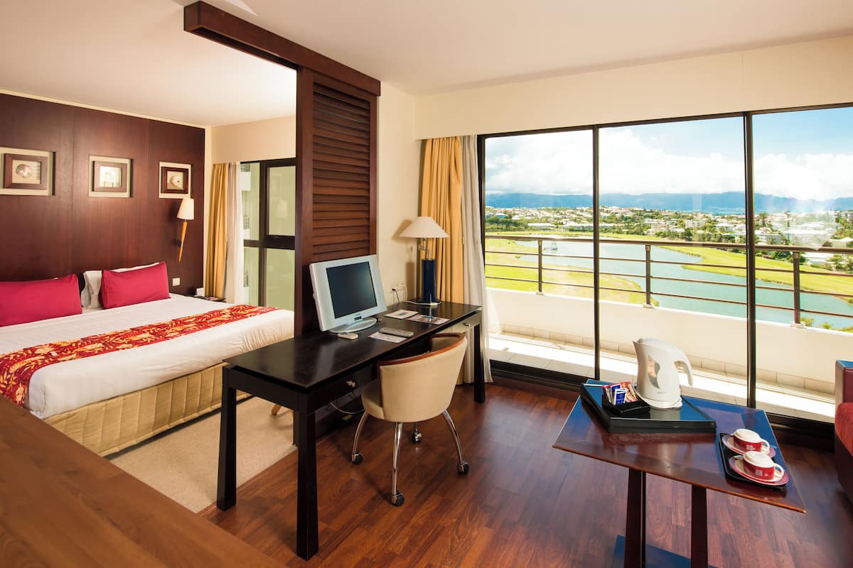 Quarto de hotel com cama, escrivaninha e uma grande janela com vista para um campo de golfe e um corpo d'água.