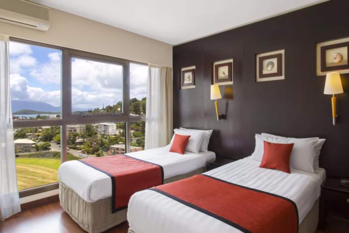 Quarto de hotel com duas camas, uma janela grande com vista para a cidade e luminárias fixadas na parede.
