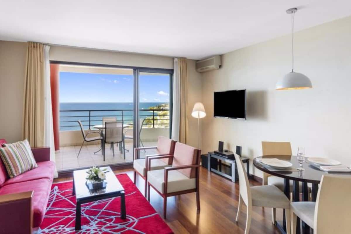 Quarto de hotel com varanda, sofá, mesa de jantar e televisão.