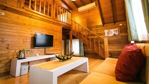 Interior de uma cabana de madeira com televisão, móveis brancos, escada e um travesseiro vermelho.