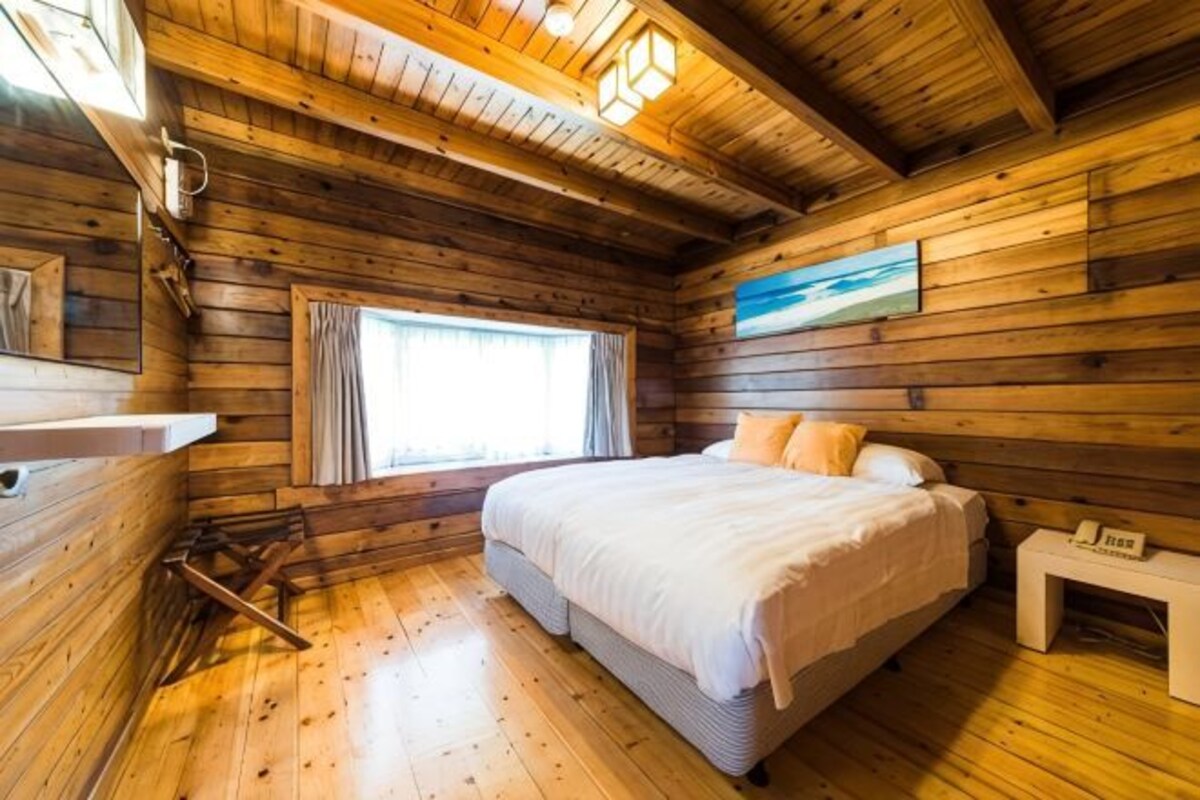 Quarto de cabana de madeira com uma cama, janela com cortinas, um quadro na parede e uma pequena mesa de cabeceira.