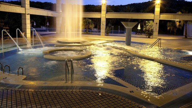 Piscina ao ar livre aquecida com diversos atrativos, incluindo uma fonte central e hidromassagens ao redor, cercada por uma área coberta com assentos e plantas em vasos.