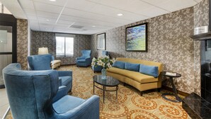 Um lobby de hotel com carpete estampado, poltronas azuis, um sofá amarelo e uma pequena mesa com um vaso de flores.