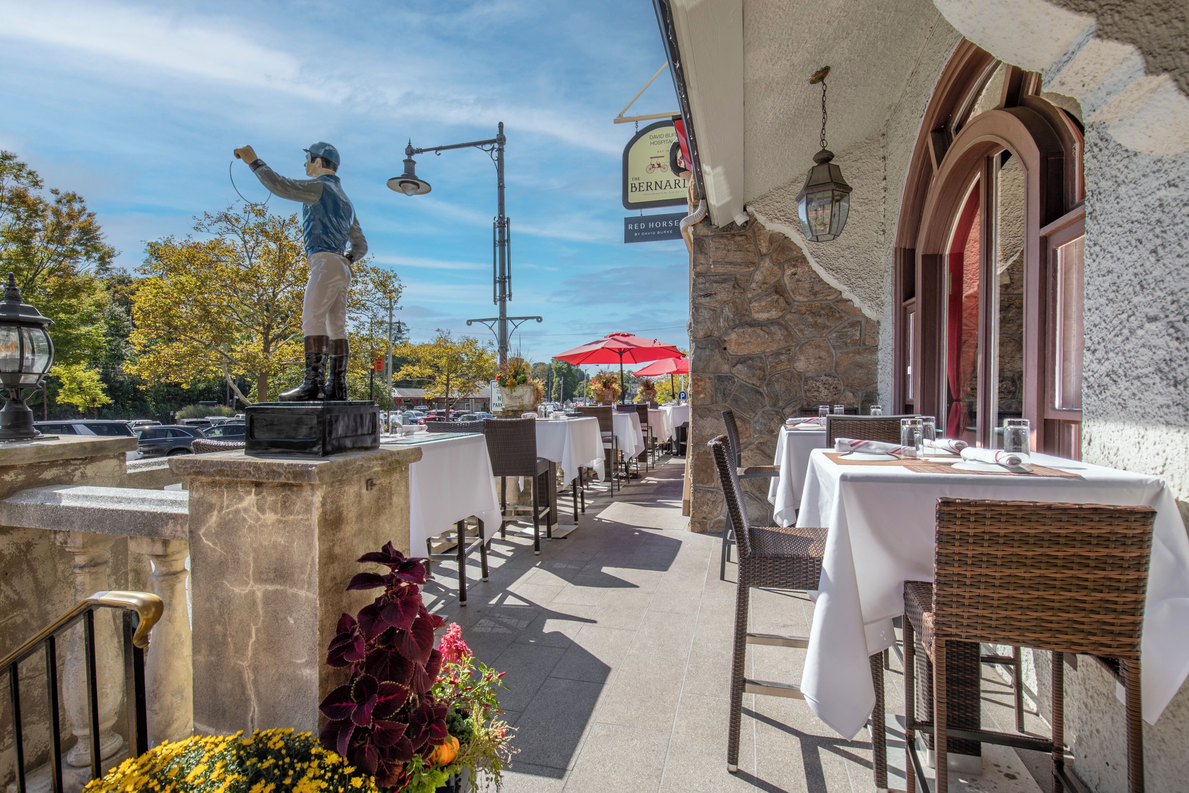 Uma área de refeições ao ar livre com mesas e cadeiras, uma estátua e um letreiro para o restaurante 'The Bernardi Red Horse'.