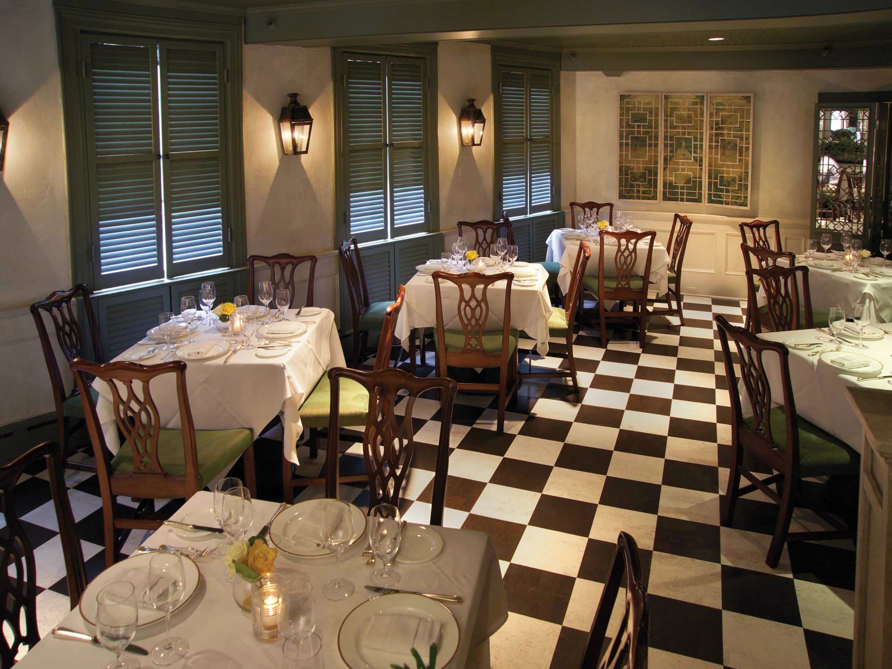 Uma sala de jantar com chão xadrez, cadeiras de madeira e mesas preparadas para refeições.