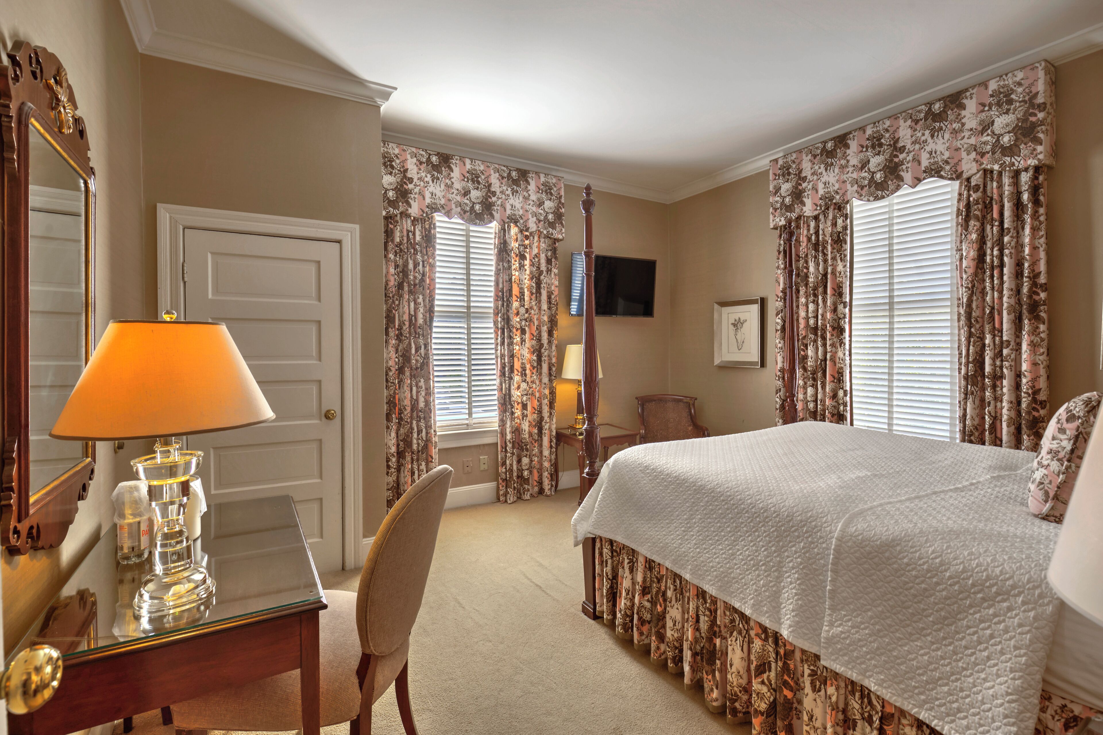 Quarto de hotel com uma cama, uma escrivaninha com candeeiro, uma cadeira, uma televisão e cortinas com padrão floral.