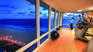 Academia com amplas janelas de frente para o oceano, equipamentos de ginástica e piso de madeira.