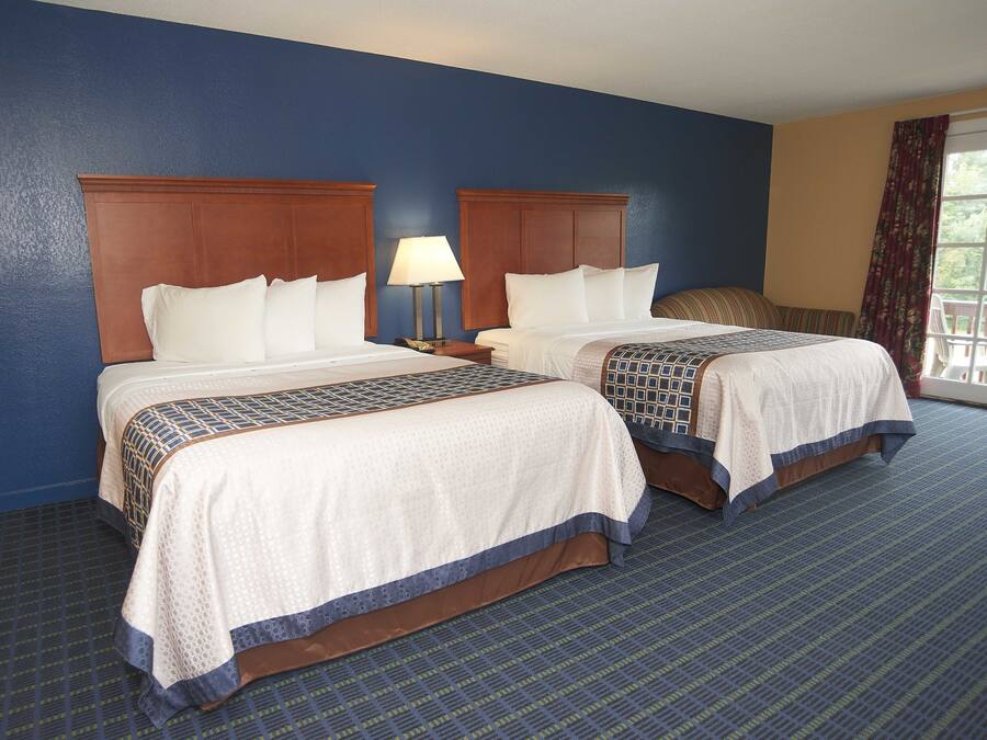 Quarto de hotel com duas camas, mesa de cabeceira, abajur e janela com cortinas.