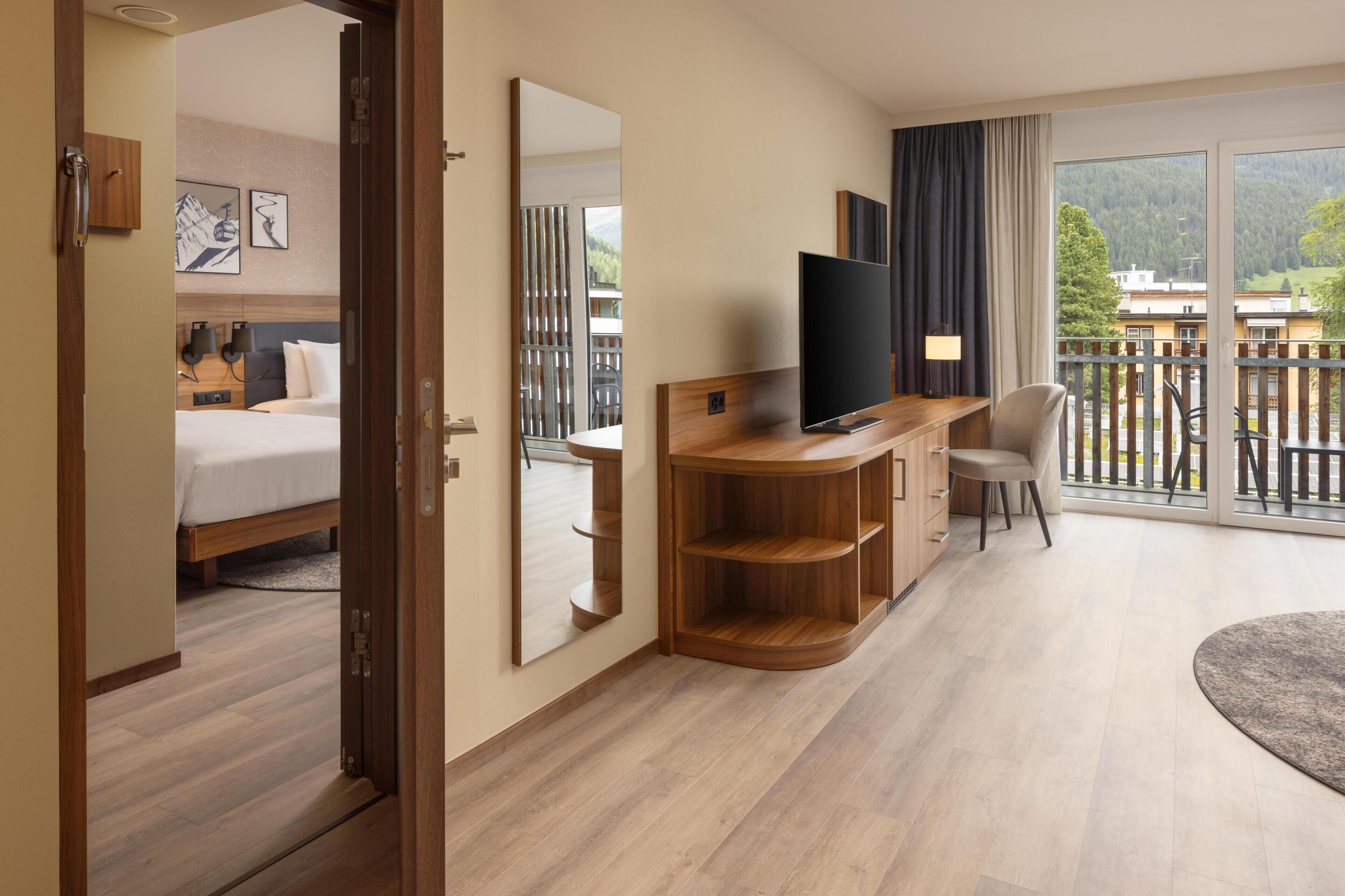 Quarto de hotel com cama, televisão, secretária com cadeira, espelho grande e varanda com vista para as montanhas.