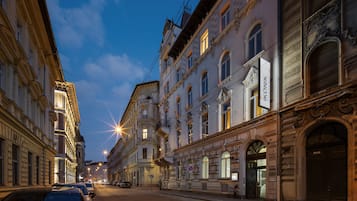 Vista da rua de um edifício histórico com arquitetura elaborada, céu claro e carros estacionados.