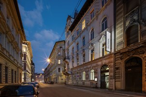 Vista da rua de um edifício histórico com arquitetura elaborada, céu claro e carros estacionados.