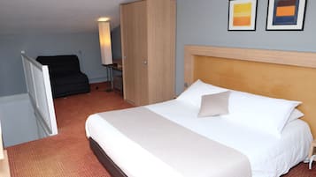 Quarto de hotel com uma cama grande, um guarda-roupa, uma escrivaninha e duas obras de arte emolduradas na parede.