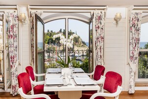 Uma sala de jantar com uma mesa posta para quatro pessoas, rodeada por cadeiras vermelhas, com vista para uma paisagem costeira através de grandes janelas.