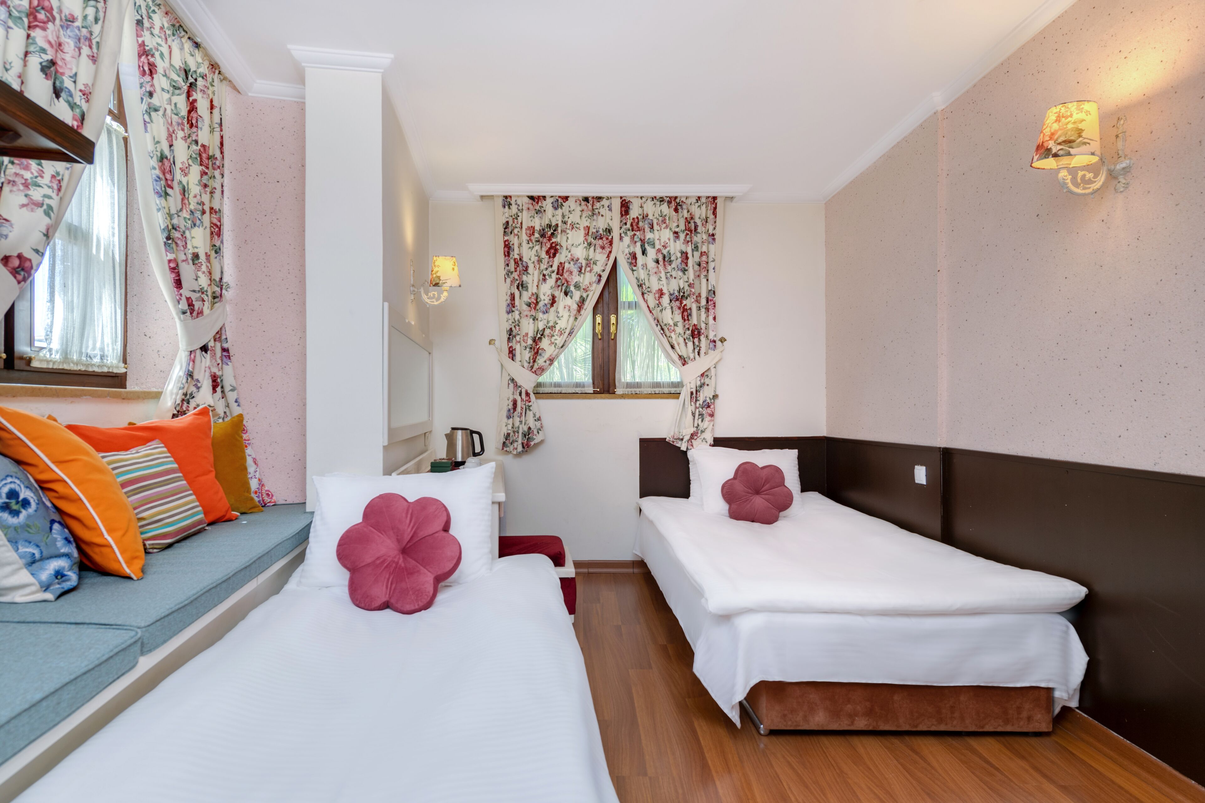 Quarto de hotel com duas camas, um sofá, cortinas florais, uma luminária na parede e uma janela com cortinas.
