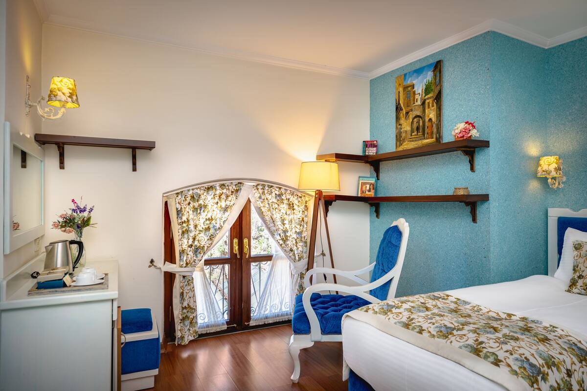 Um quarto com cama, uma cadeira, escrivaninha com luminária e prateleira com um quadro e um vaso.
