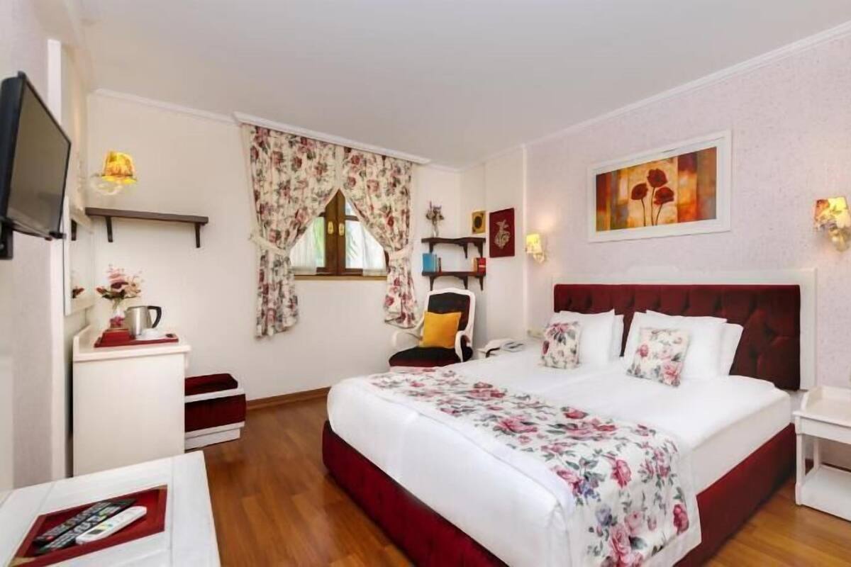Quarto de hotel com uma cama grande, uma escrivaninha, uma cadeira, uma televisão e uma janela com cortinas florais.