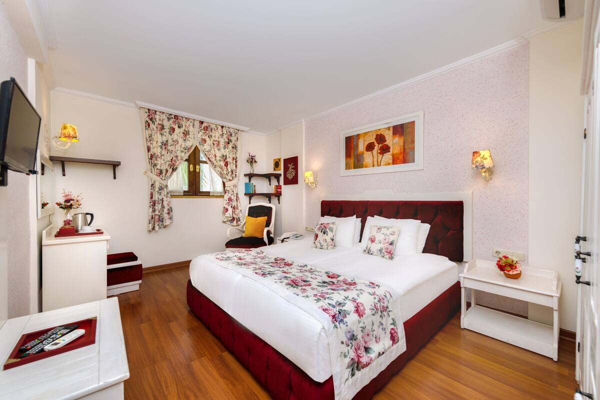 Quarto de hotel com uma cama grande, uma mesa, uma cadeira, uma televisão e uma janela com cortinas florais.