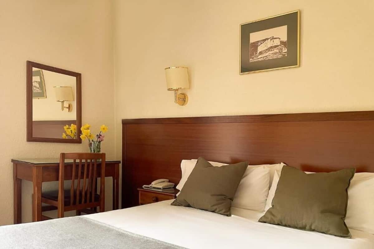 Quarto de hotel com uma cama grande, uma mesa de cabeceira de madeira, uma escrivaninha com cadeira, uma luminária e um quadro na parede.