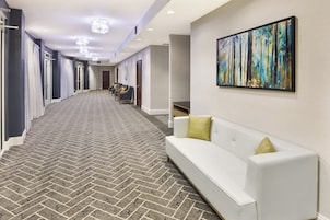 Corredor de hotel com carpete estampado, um sofá branco com almofadas e um quadro na parede.