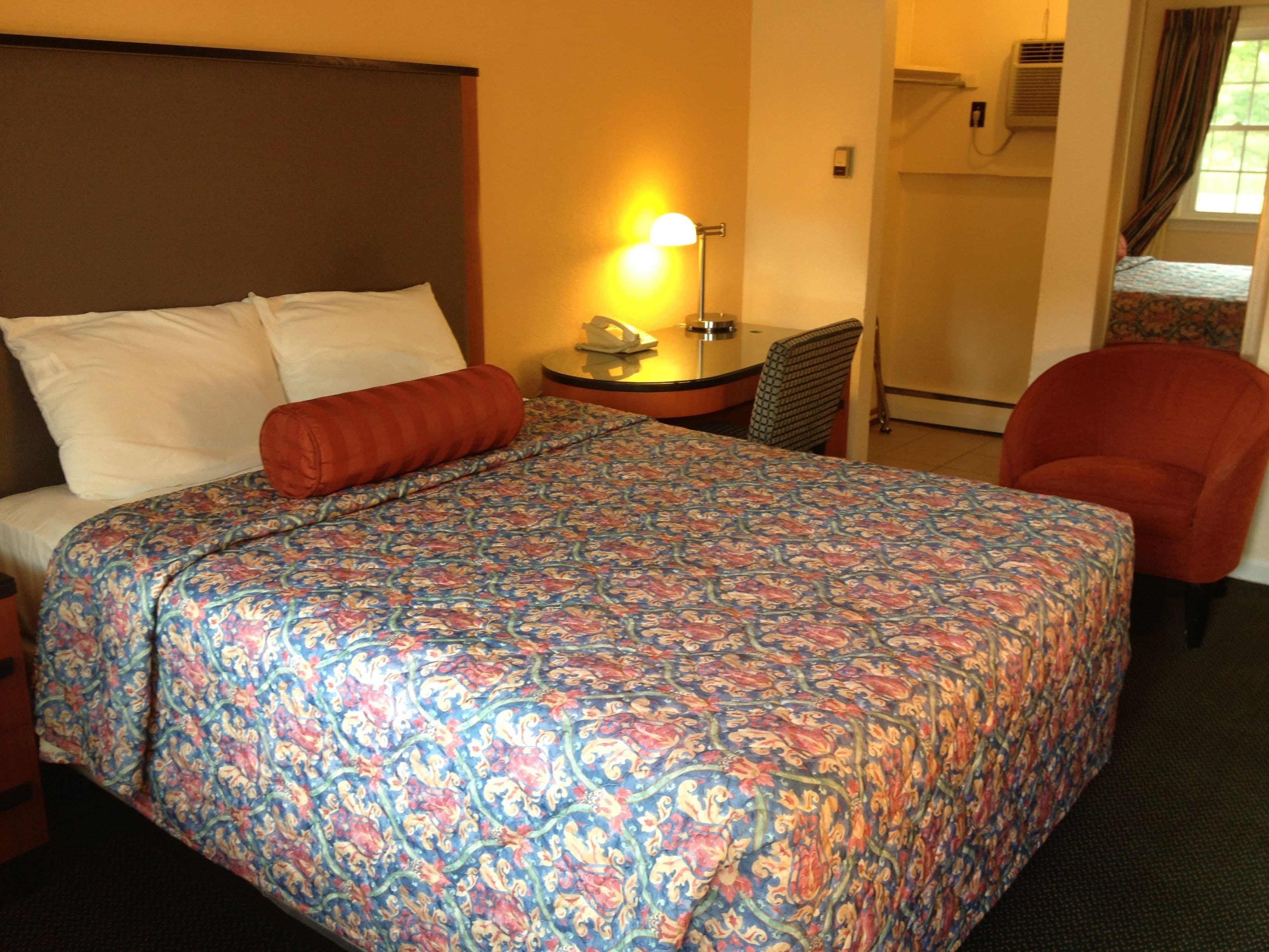 Quarto de hotel com cama, mesa de cabeceira, luminária, cadeira e ar condicionado.