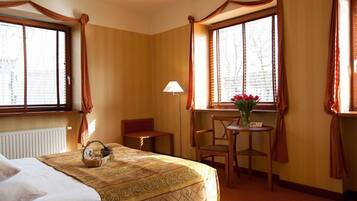 Quarto de hotel com cama, uma mesinha de cabeceira com abajur, uma cadeira e uma janela com cortinas.