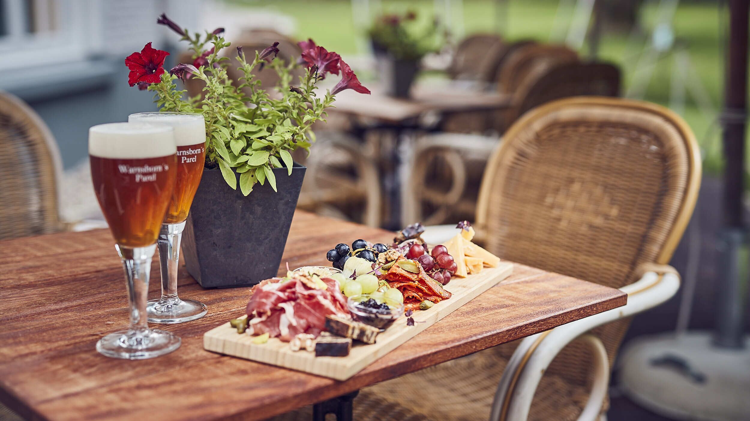 Uma mesa de madeira com um tabuleiro de frios, copos de cerveja e um vaso com planta.