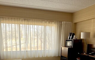 Quarto com uma janela grande coberta por cortinas finas, máquina de café, luminária e micro-ondas em uma bancada de madeira.