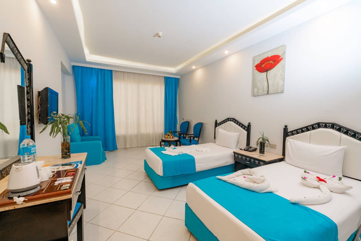 Quarto de hotel com duas camas, uma escrivaninha, uma cadeira, uma mesa pequena e uma janela grande com cortinas.