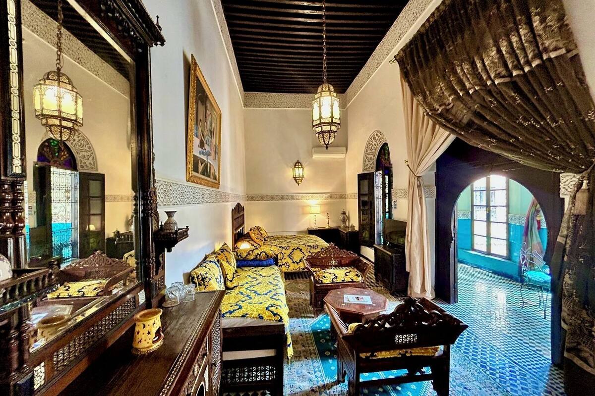 Quarto tradicional marroquino com cama, mesa de madeira, espelho e vista para o exterior.