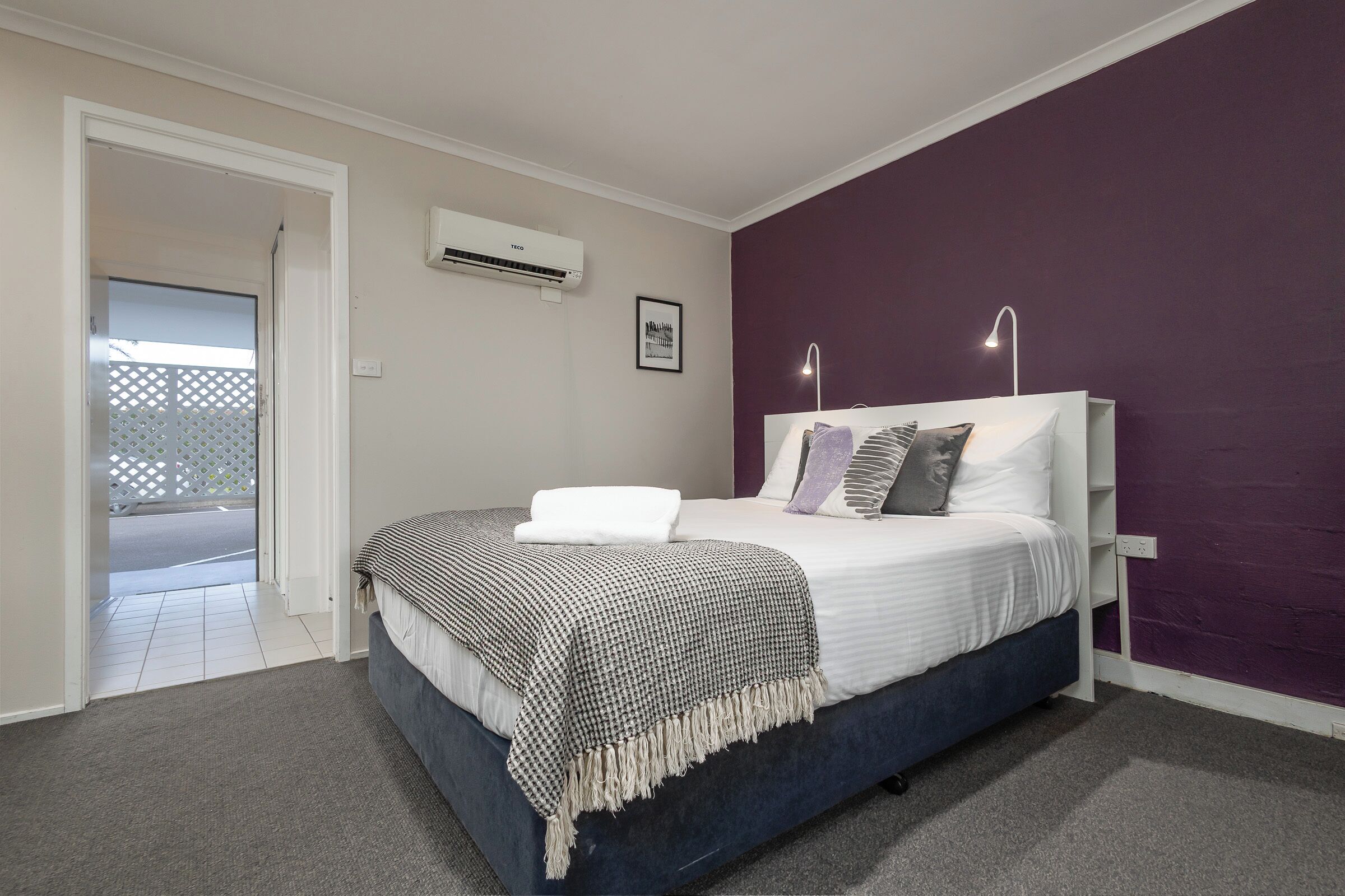 Quarto com cama, ar condicionado branco, porta que dá para uma varanda e parede roxa.