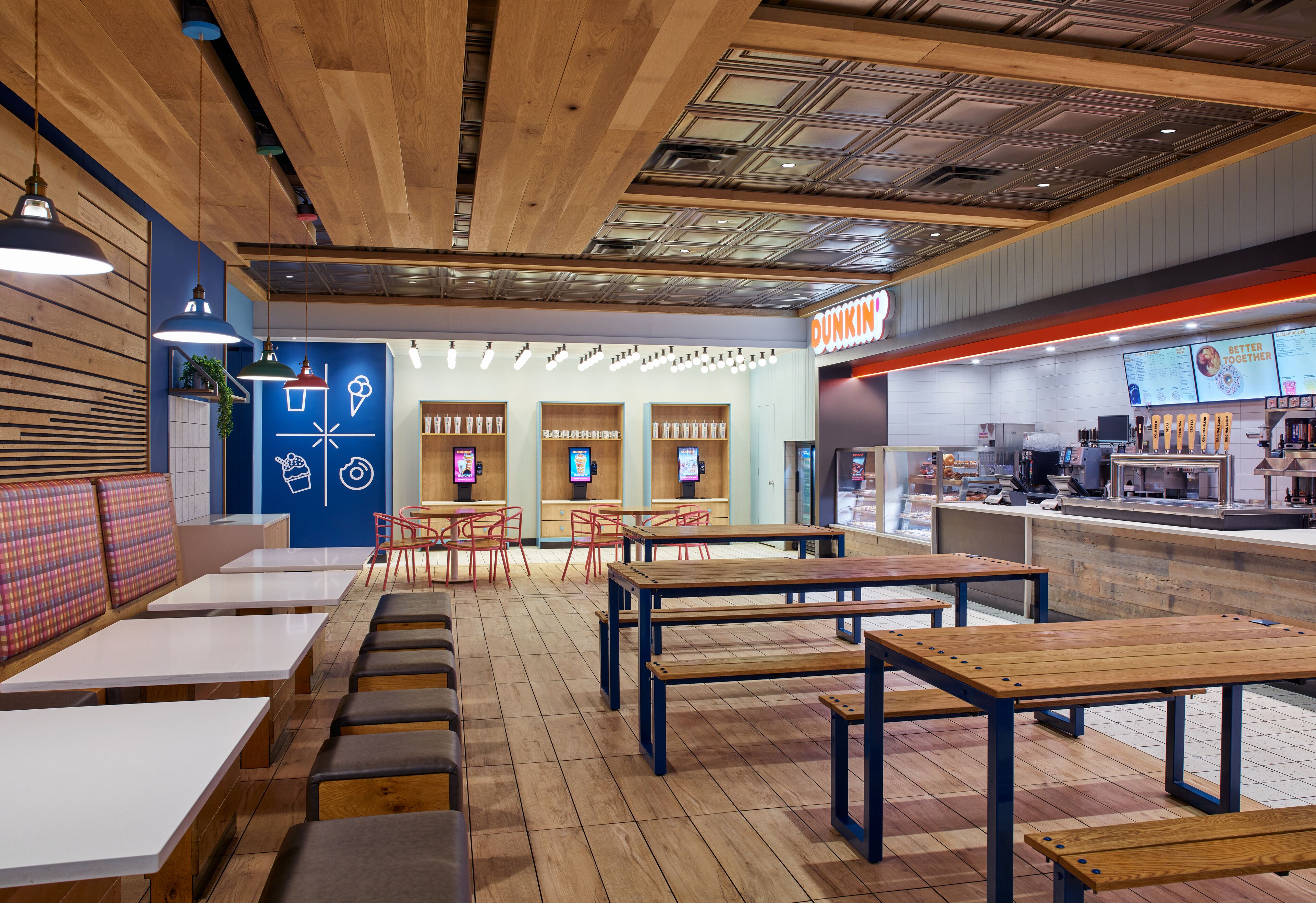Decoração moderna com mesas e bancos de madeira, um balcão da Dunkin' Donuts e uma parede azul com ícones.