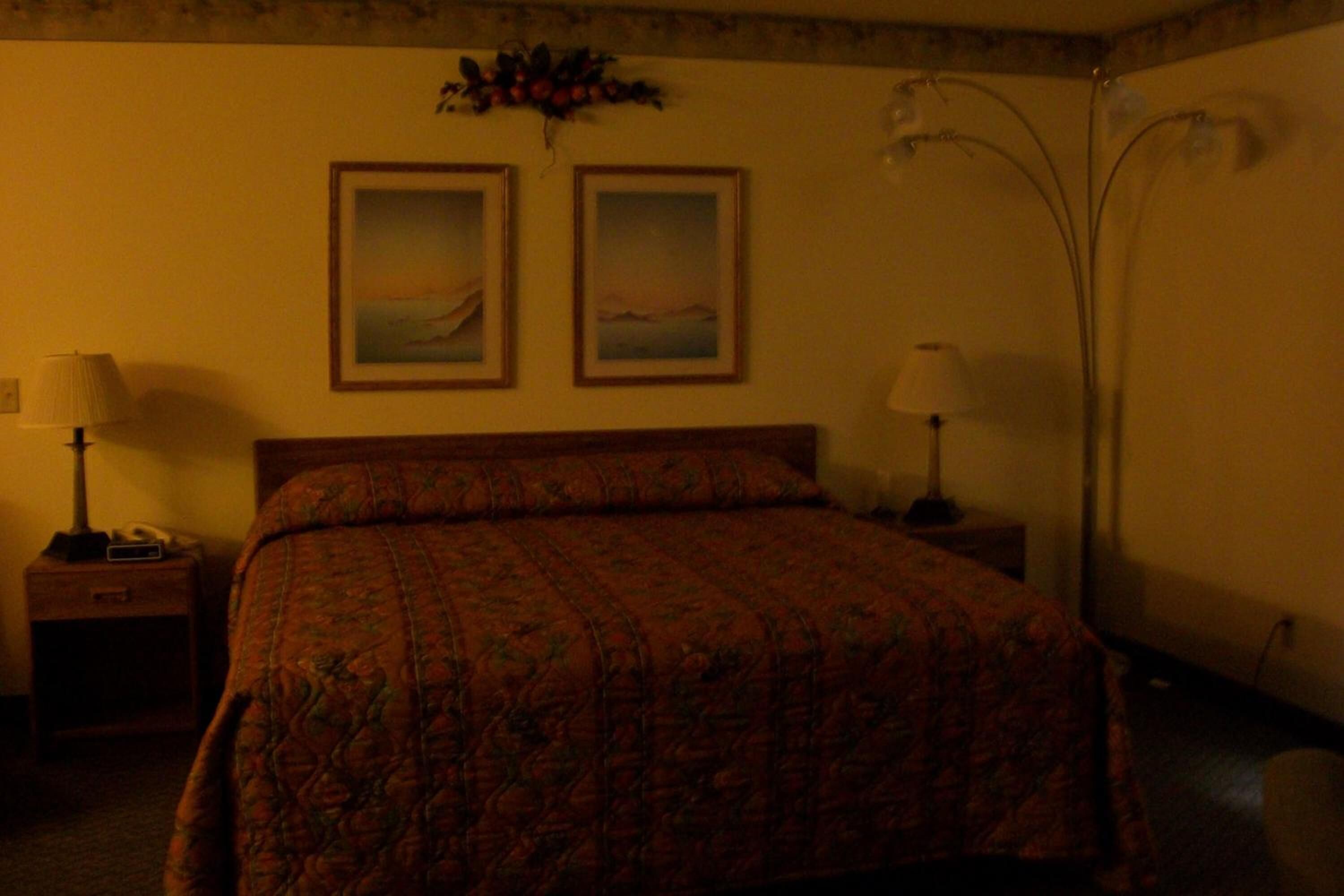 Quarto de hotel com iluminação suave, uma cama, duas molduras na parede, uma mesa de cabeceira com abajur e um candeeiro de chão alto.