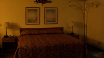 Quarto de hotel com iluminação suave, uma cama, duas molduras na parede, uma mesa de cabeceira com abajur e um candeeiro de chão alto.