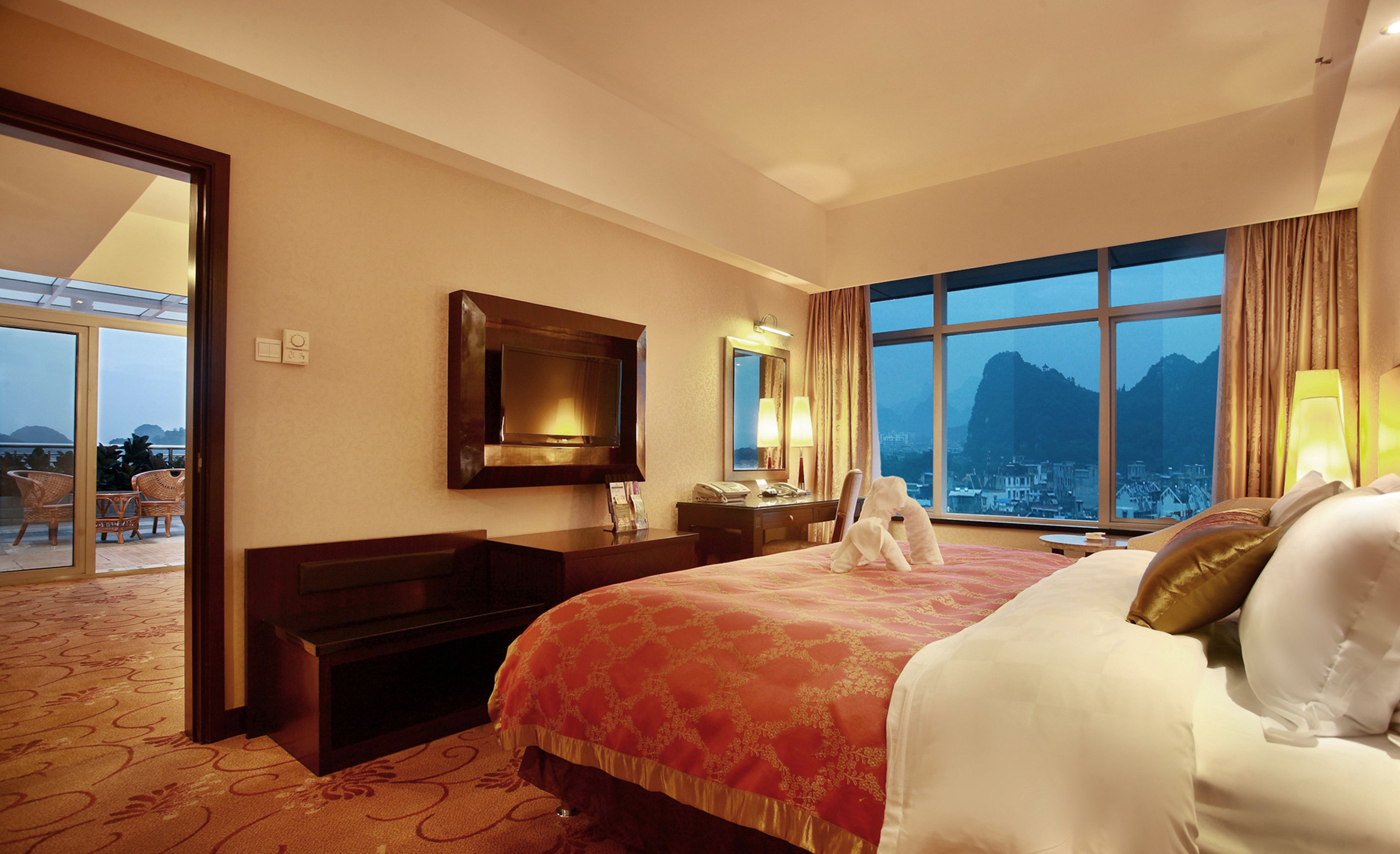 Quarto de hotel com uma cama grande, uma escrivaninha, televisão e vista para uma montanha e paisagem urbana.