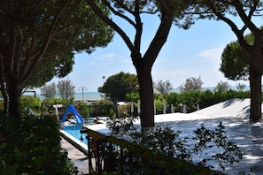 Área de piscina com um escorregador azul, rodeada por árvores e vista para o mar.