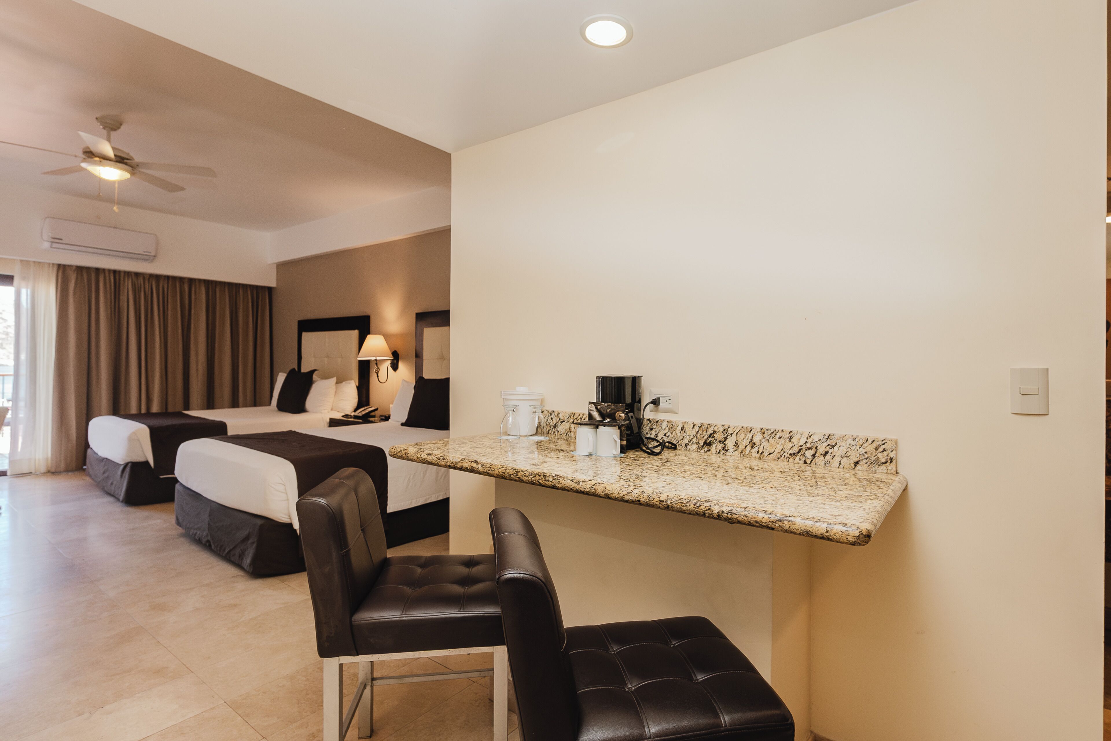 Quarto de hotel com cama, duas cadeiras, uma mesa pequena, ventoinha no teto, aparelho de ar condicionado e área de cozinha com bancada.