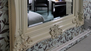 Um espelho com moldura elaborada reflete um quarto com uma cadeira, uma mesa pequena e cortinas.