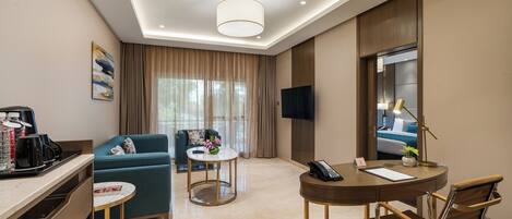 Quarto de hotel com sofá, mesa redonda pequena, escrivaninha com cadeira, televisão e luminária no teto.