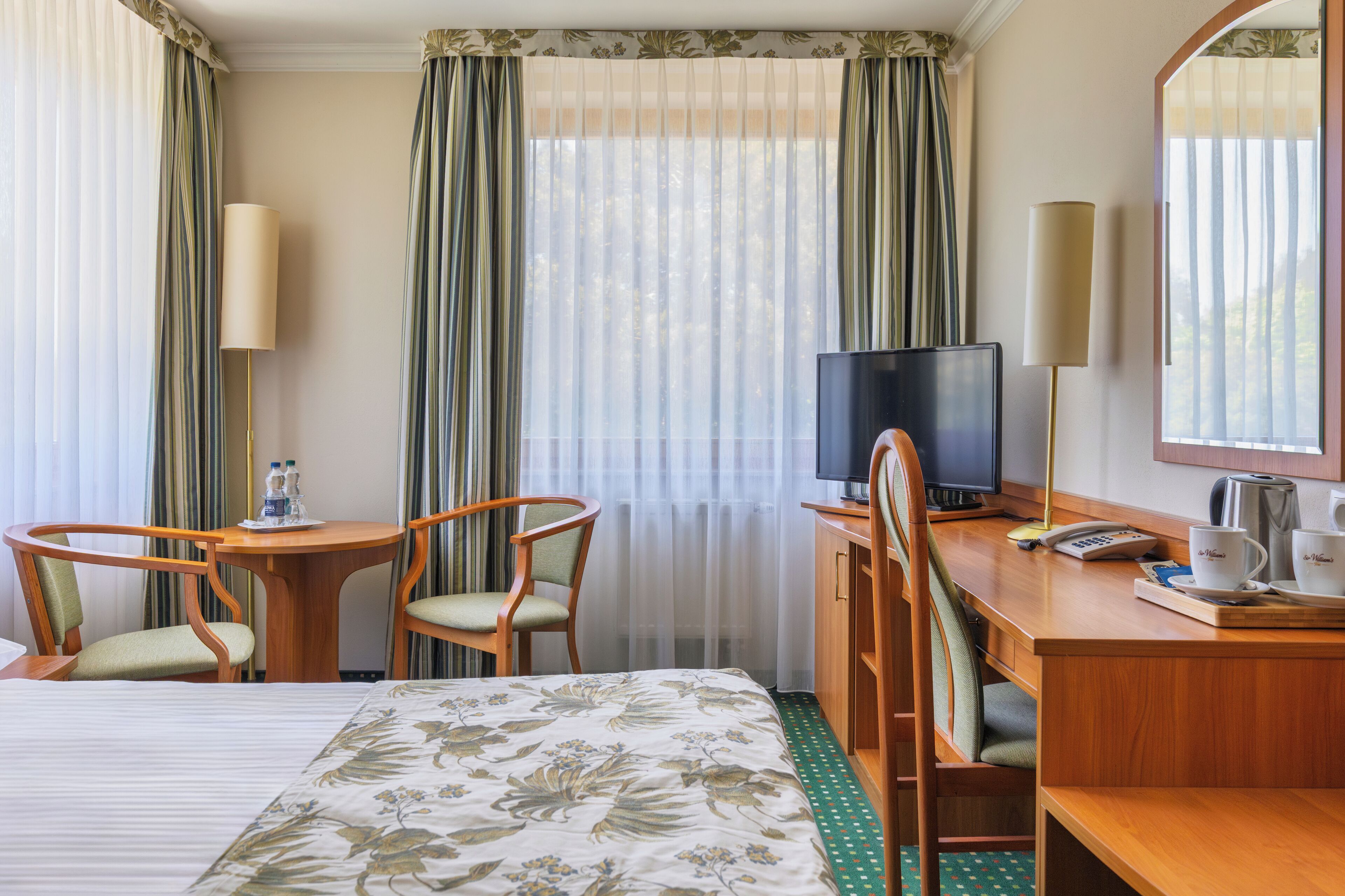 Quarto de hotel com uma cama, duas cadeiras, uma escrivaninha com televisão, telefone e espelho.