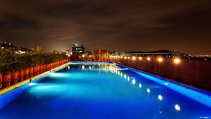 Piscina iluminada à noite, com deck de madeira e vasos de plantas na borda.