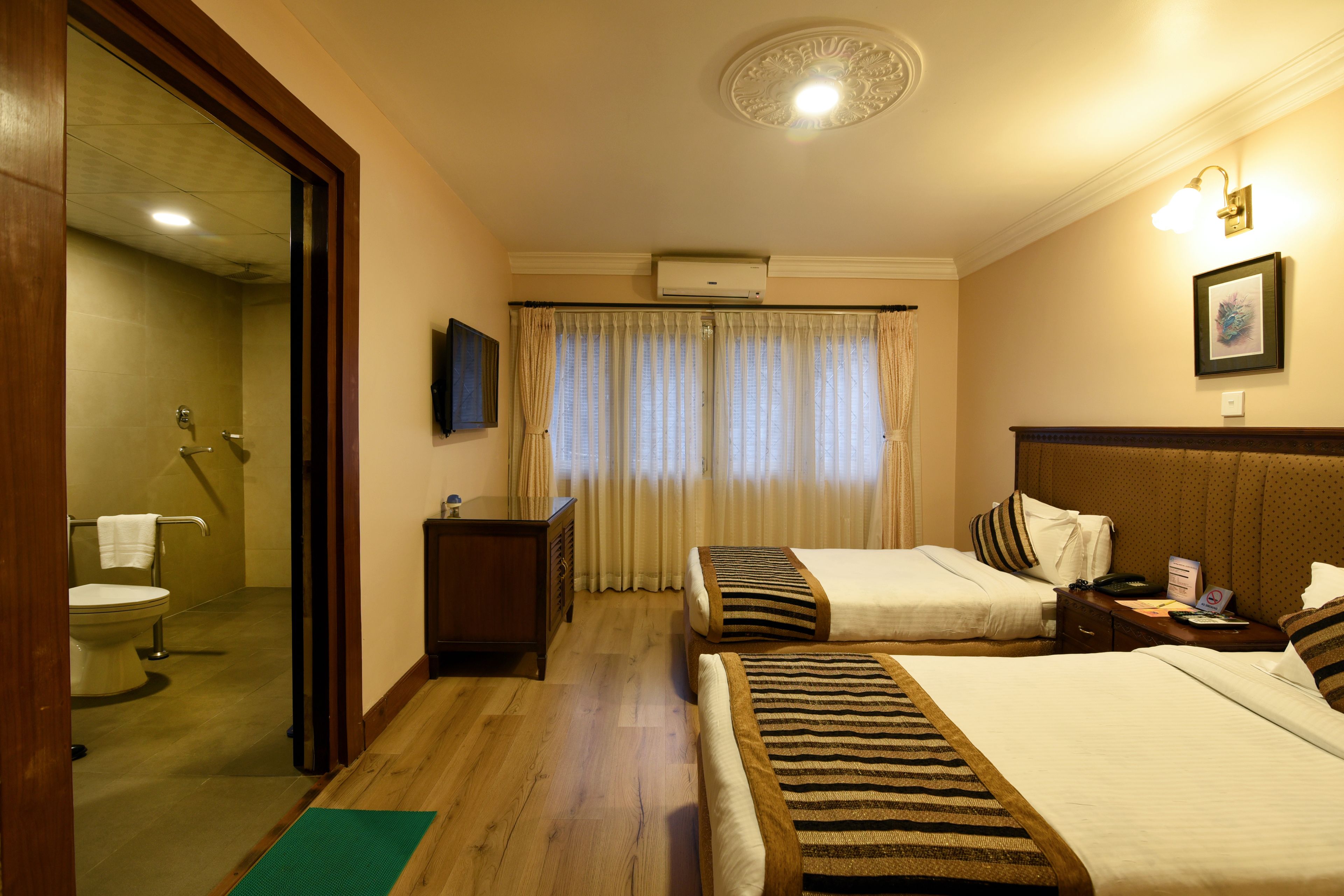Quarto de hotel com duas camas, chão de madeira, banheiro com vaso sanitário e pia, e um espelho grande.