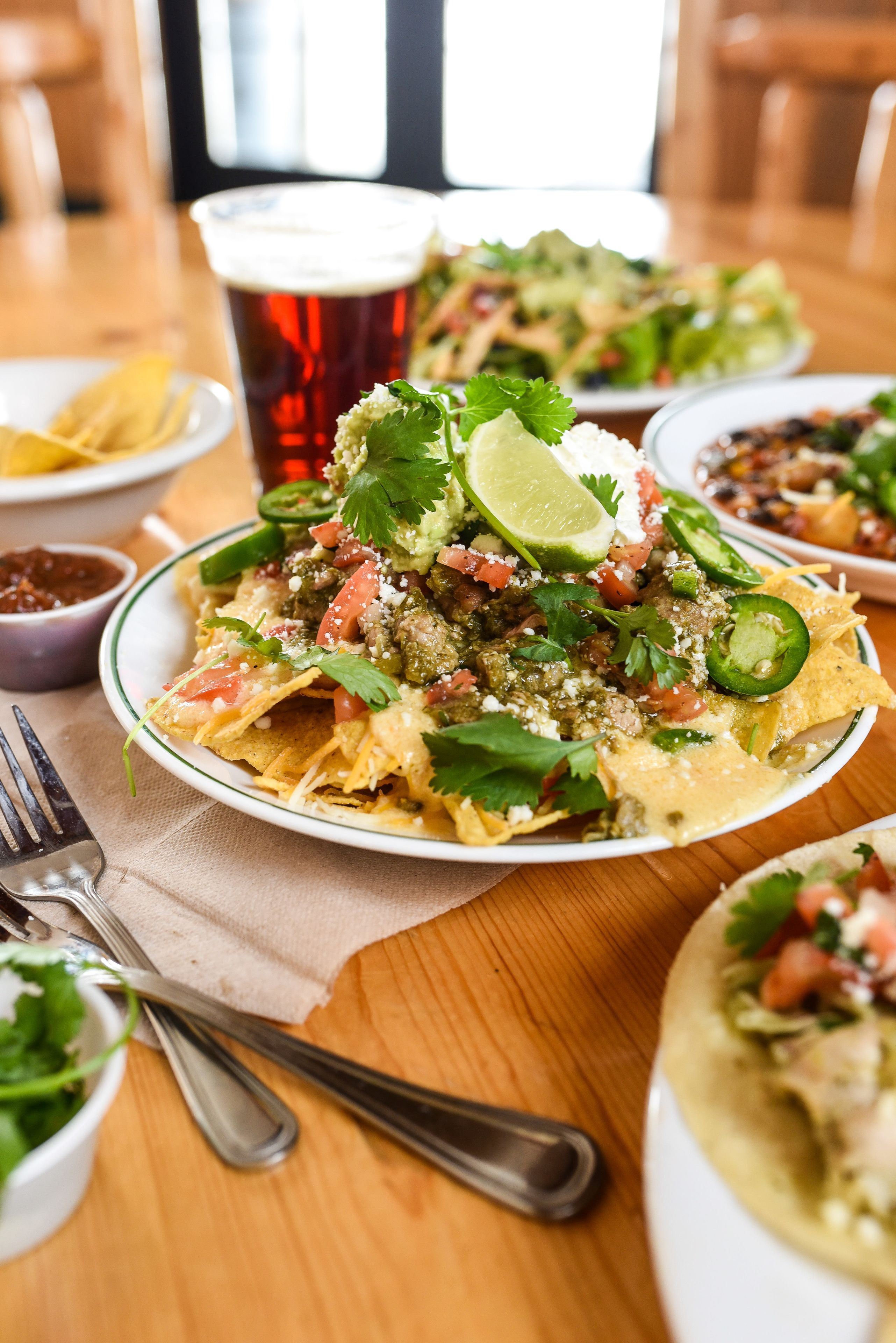 Um prato de nachos coberto com diversos ingredientes, um copo de cerveja e uma tigela de molho de tomate sobre uma mesa de madeira.
