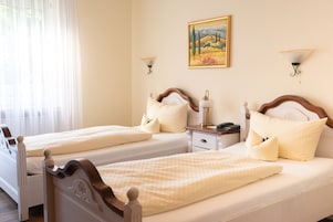 Quarto de hotel com duas camas de solteiro, mesa de cabeceira com abajur, um quadro na parede e janela com cortinas leves.