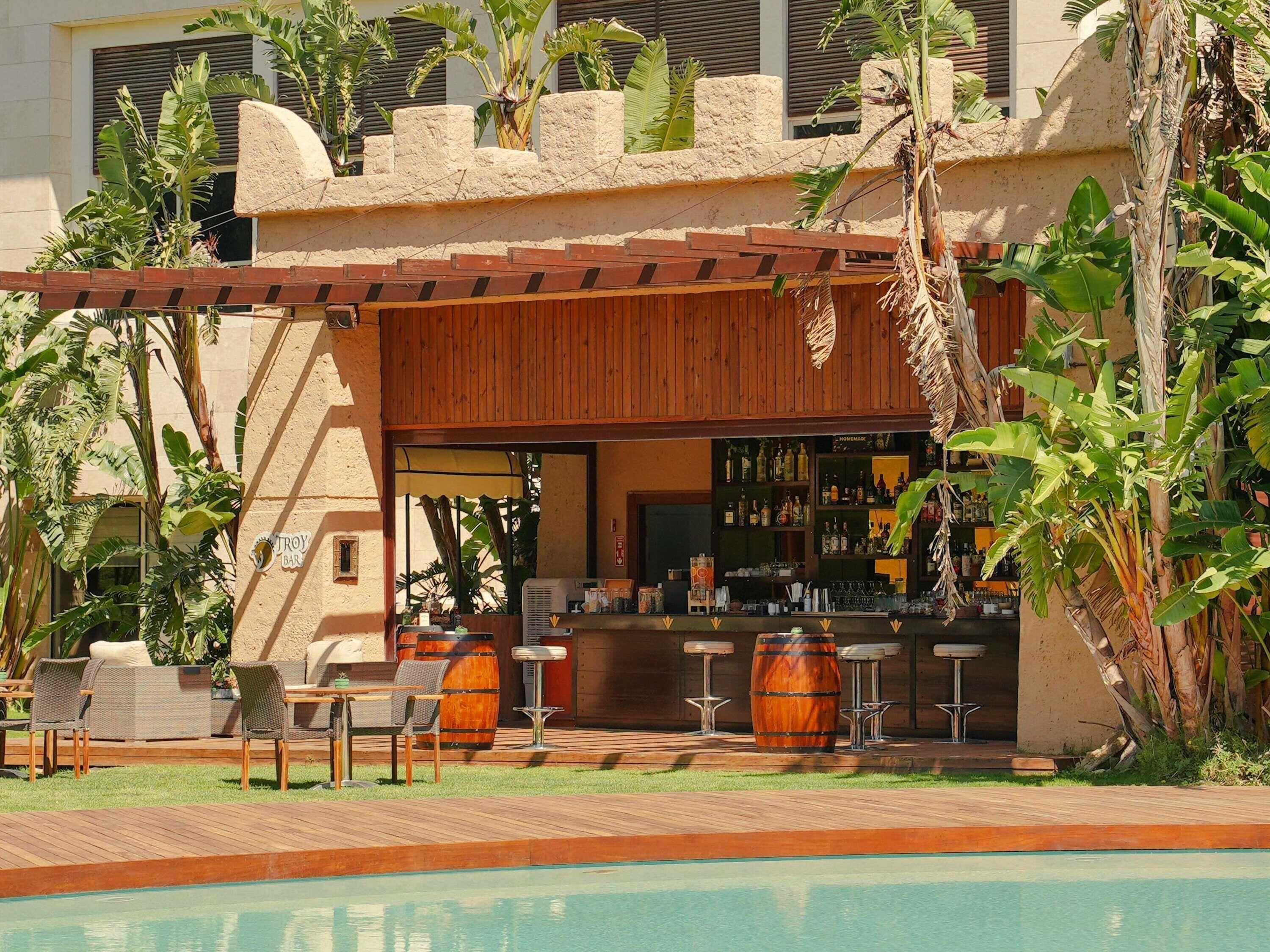 Uma área à beira da piscina com bar, assentos e plantas tropicais.