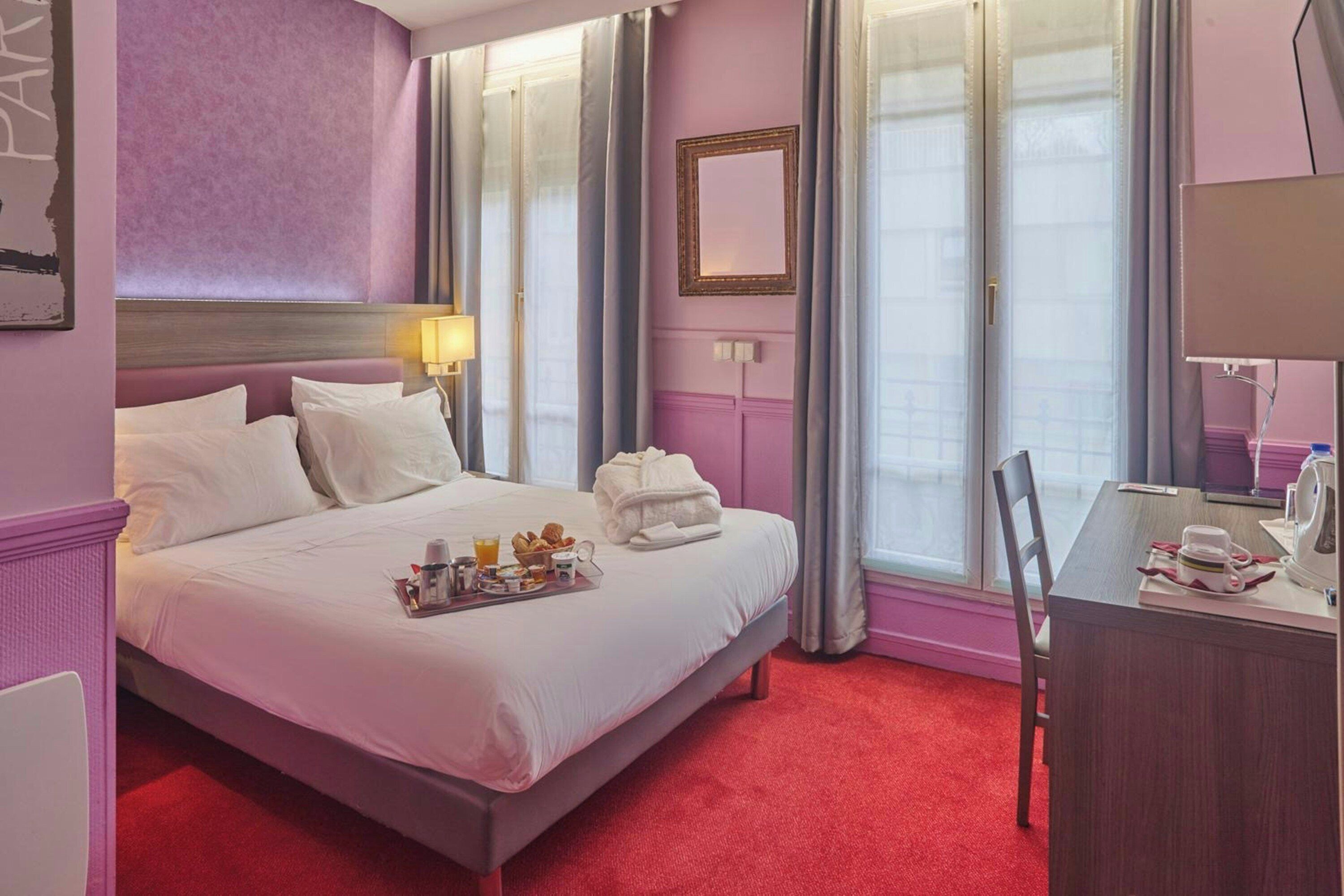 Quarto de hotel com cama, escrivaninha, cadeira, janela com cortinas e espelho fixado na parede.