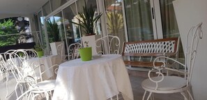 Uma área de pátio ao ar livre com cadeiras e mesas de metal branco, um banco de madeira e plantas em vasos.