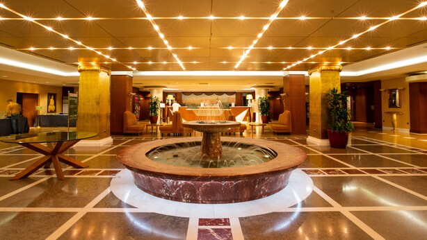 Um hall amplo com uma fonte central, balcão de recepção e áreas de estar.